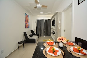 Uma sala de jantar com mesa posta para quatro pessoas, um ventilador de teto, um sofá e uma televisão ao fundo.