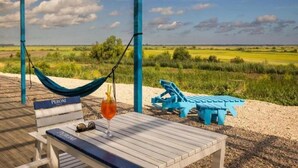 Um deck de madeira com uma rede, uma mesa com uma bebida e uma cadeira com a etiqueta 'PERONI', com vista para um campo verde.