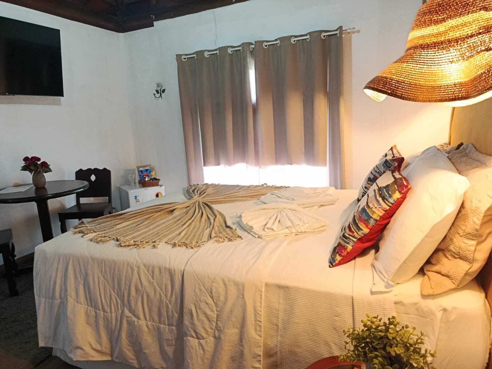 Quarto com cama, mesa com um vaso de flores, cadeira, janela com cortinas e luminária pendente.