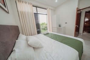 Quarto com cama, janela com cortinas, banheiro com pia e porta.
