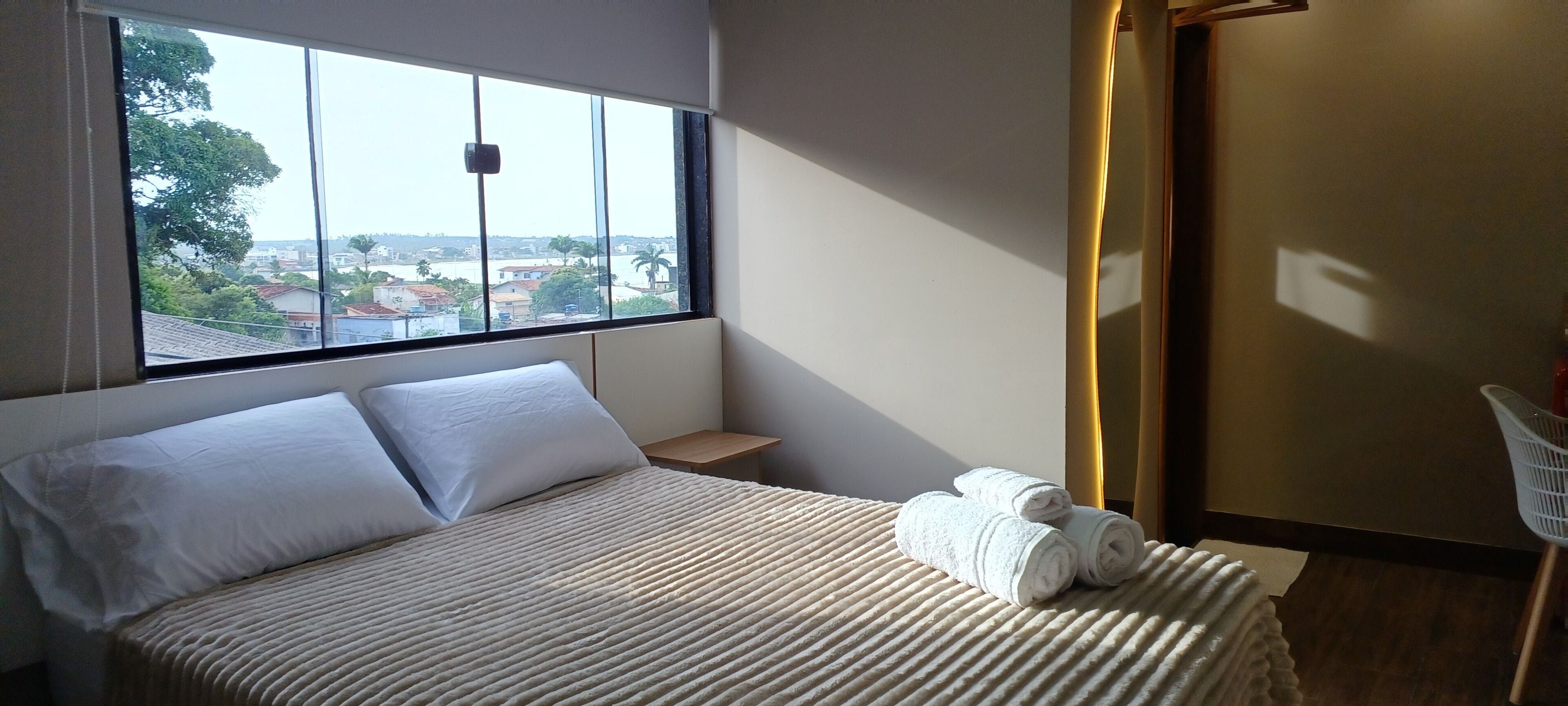 Quarto de hotel com cama, janela vista para edifícios, mesa de cabeceira pequena e toalha sobre a cama.