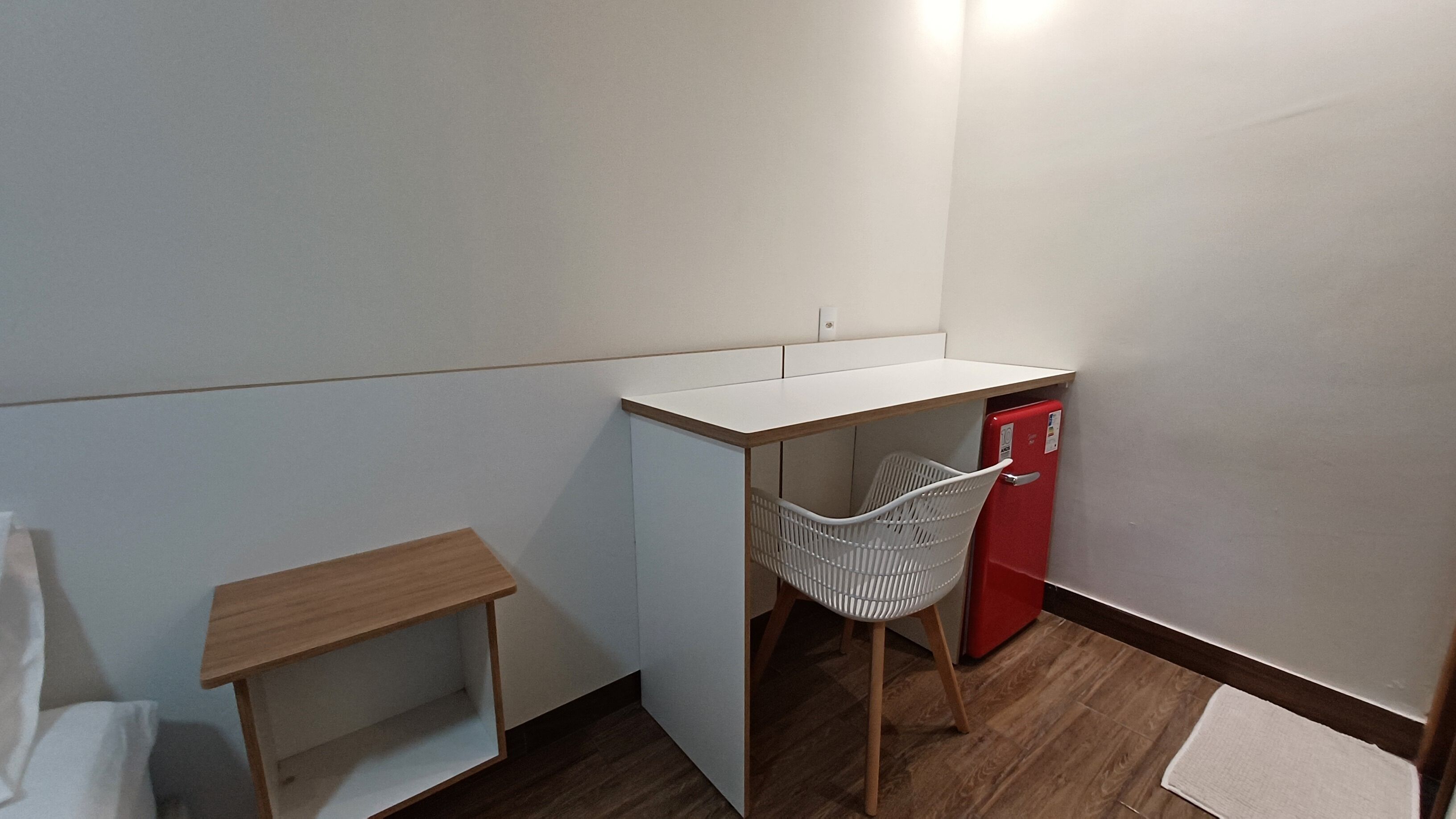 Quarto pequeno e compacto com escrivaninha, cadeira, geladeira vermelha e prateleira de madeira.