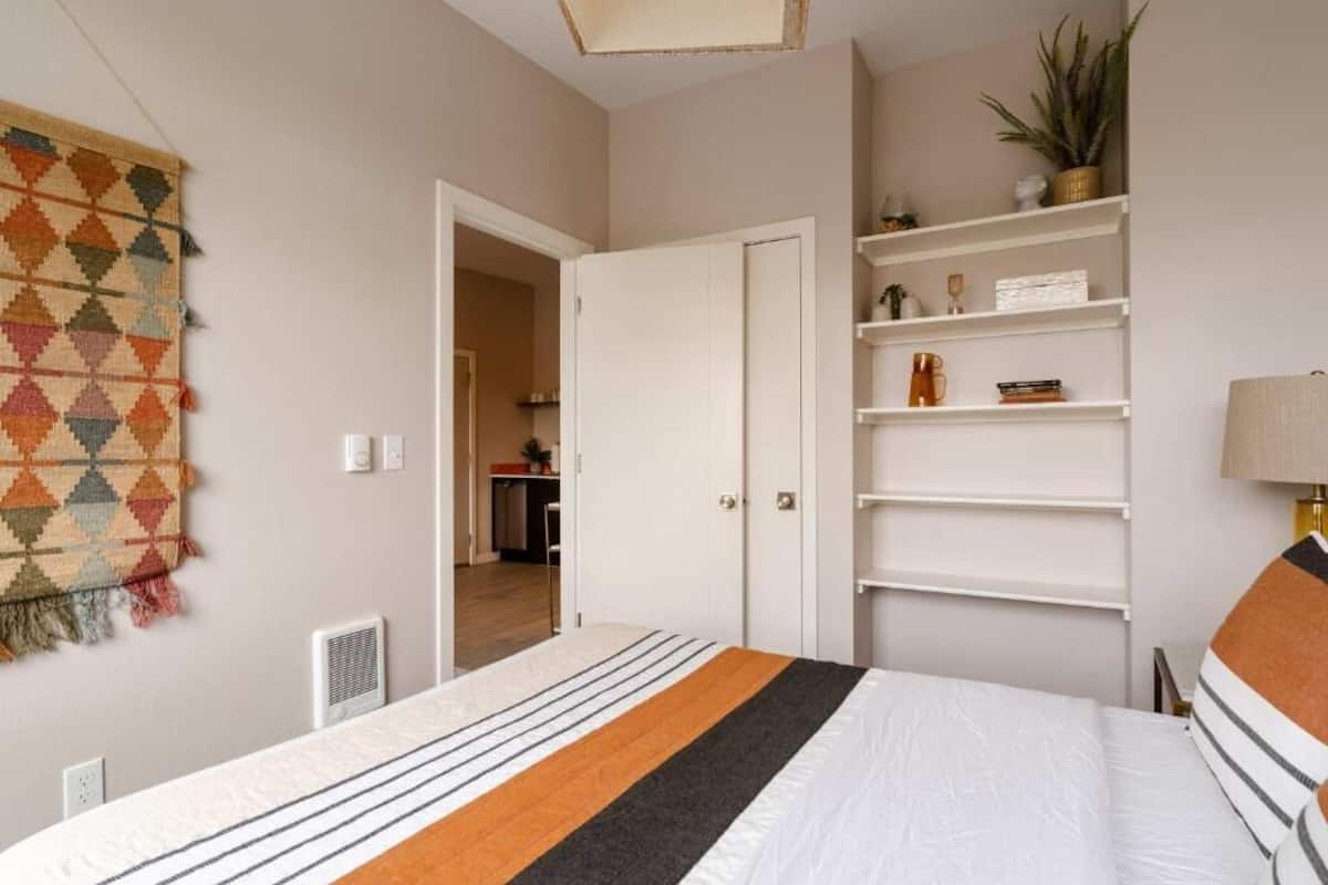 Quarto com cama, prateleira fixada na parede com objetos decorativos e porta para outro cômodo.