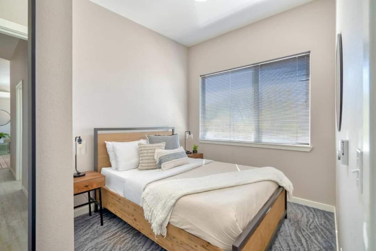 Quarto com cama, mesas de cabeceira, um criado-mudo, janela com persianas e uma porta.