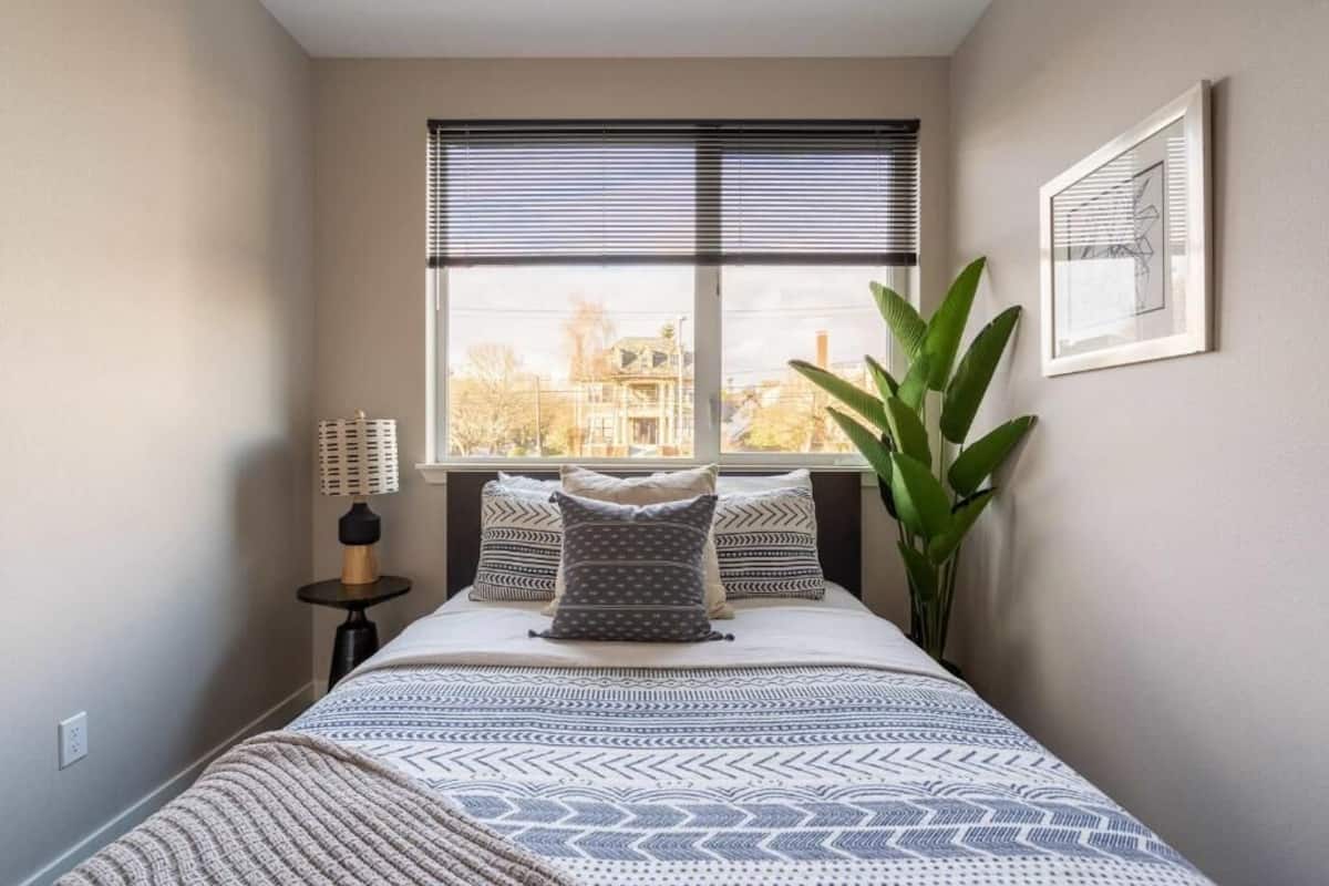 Quarto com cama, janela com persianas, mesa de cabeceira com abajur, planta em vaso e quadro na parede.