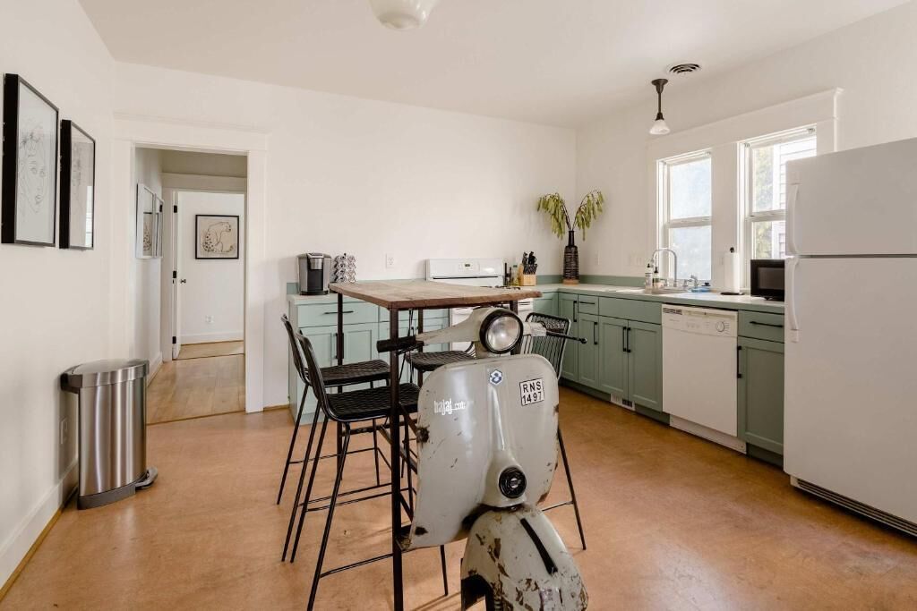 Uma cozinha com mesa de jantar, cadeiras, uma scooter antiga, um cesto de lixo e uma geladeira.