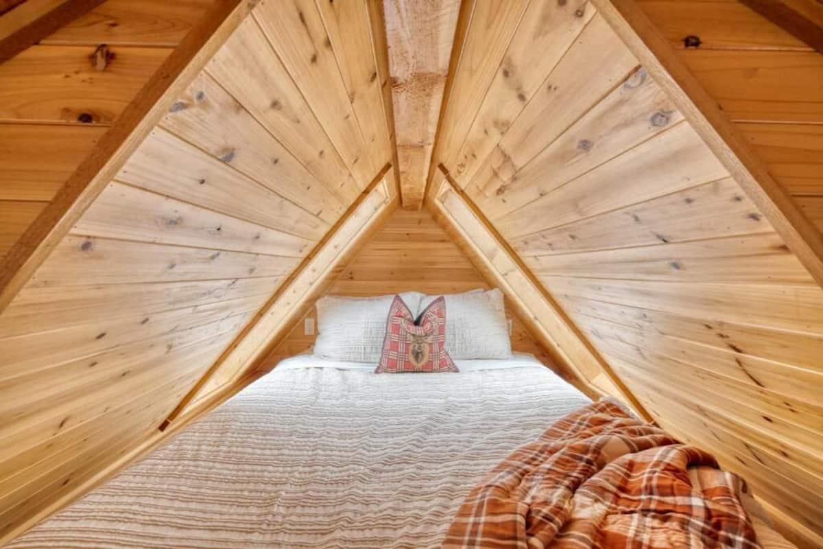 Quarto sótão triangular com paredes de madeira e cama com manta xadrez.