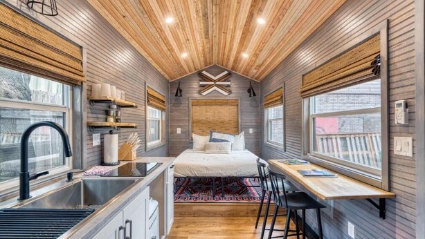 Quarto compacto com cama, pequena cozinha, área de refeições e amplas janelas.