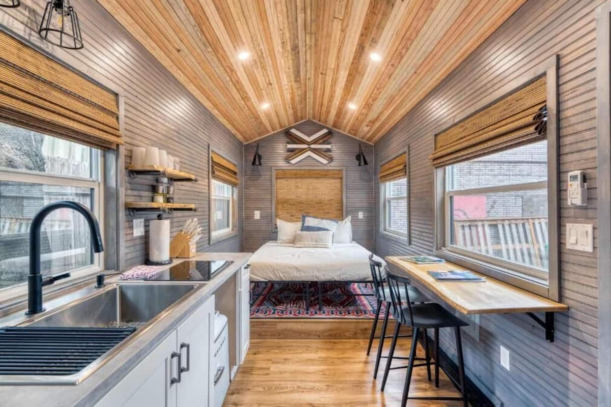 Quarto compacto com cama, pequena cozinha, área de refeições e amplas janelas.