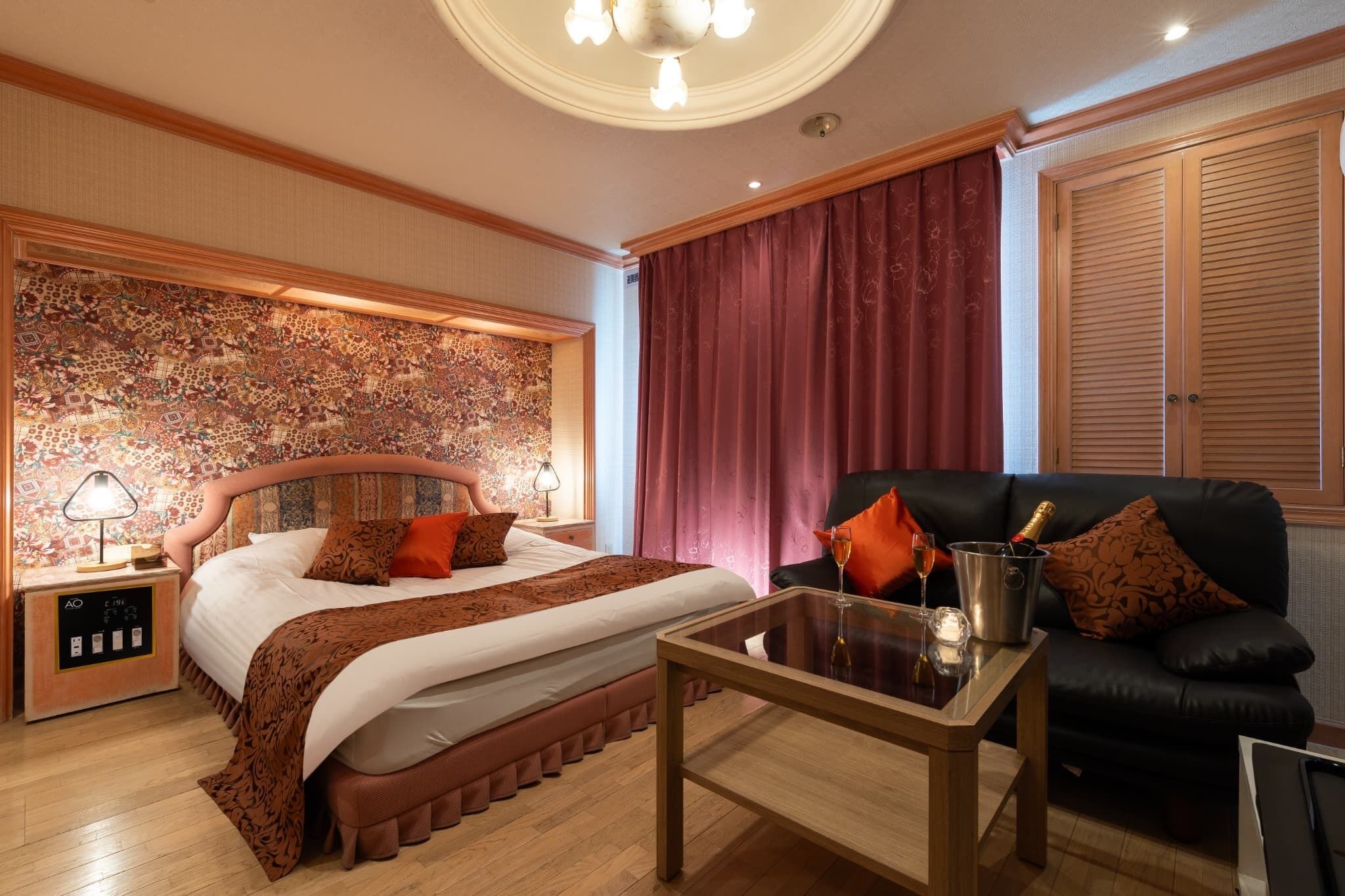 Quarto de hotel com uma cama, um sofá preto, uma mesa de vidro, uma cortina vermelha, uma cabeceira de madeira e uma parede com desenho estampado.