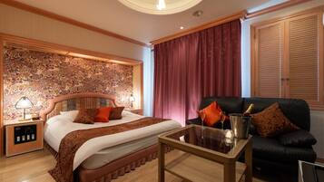 Quarto de hotel com uma cama, um sofá preto, uma mesa de vidro, uma cortina vermelha, uma cabeceira de madeira e uma parede com desenho estampado.