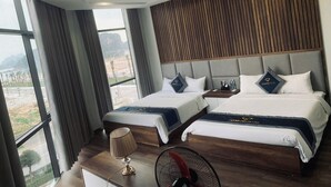 Quarto de hotel com duas camas, uma escrivaninha, abajur, ventilador e vista para o exterior.