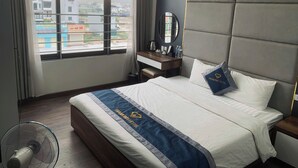 Quarto de hotel com cama, mesa de cabeceira, espelho, janela e vista para edifícios, e um ventilador.