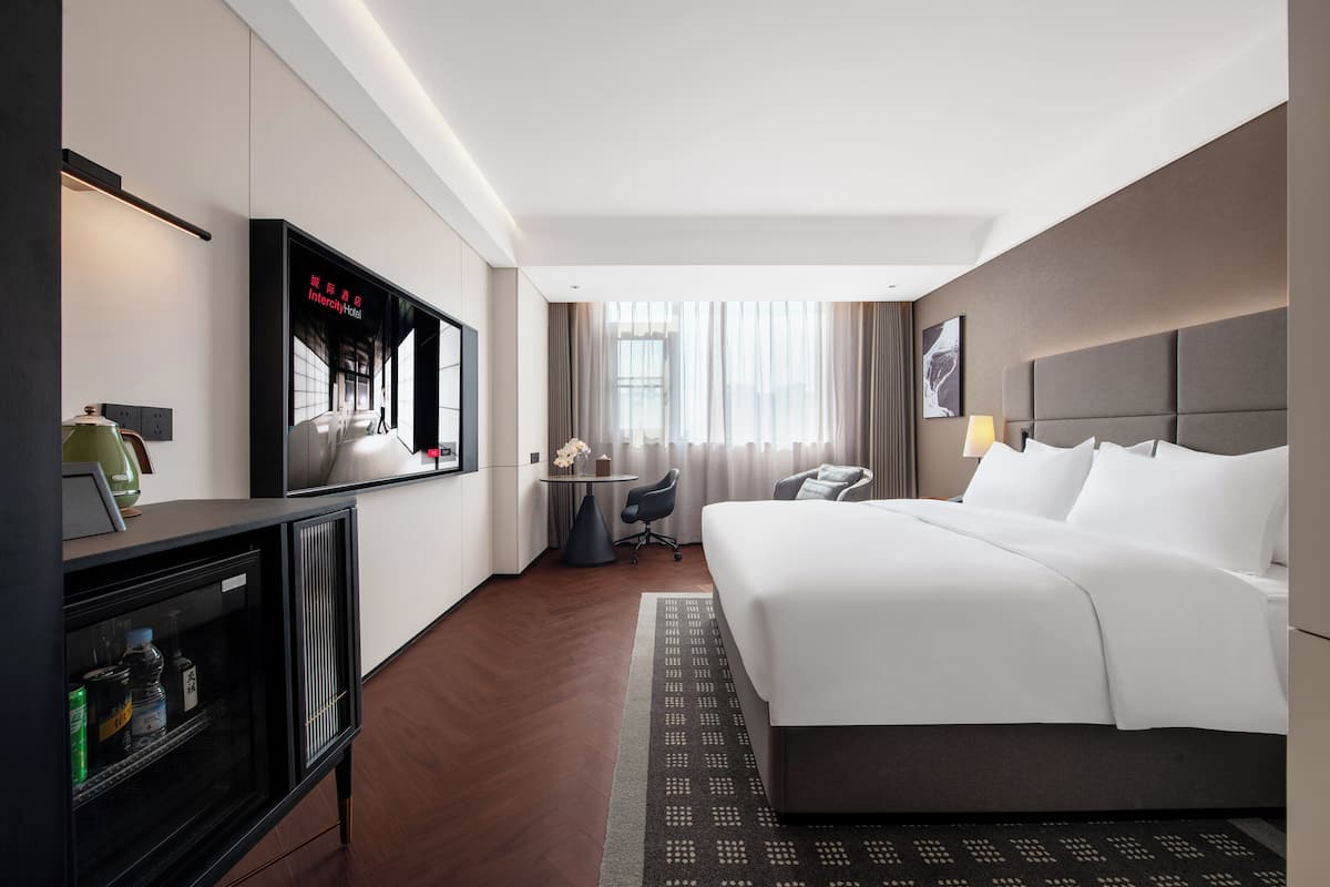 Quarto de hotel com uma cama grande, uma escrivaninha com cadeira, televisão e minibar.