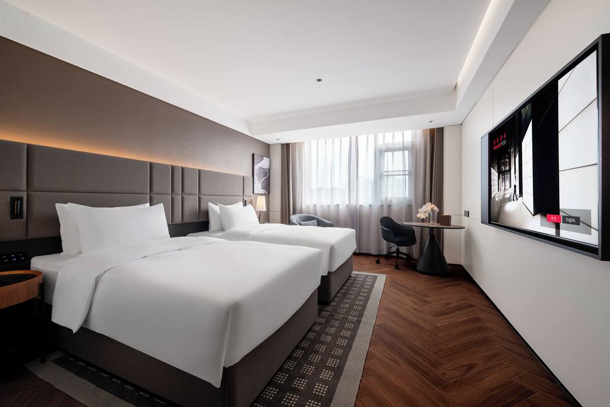 Quarto de hotel com duas camas, uma escrivaninha, uma cadeira, uma televisão e uma janela grande com cortinas.