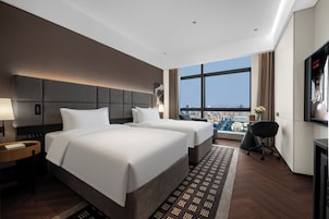 Quarto de hotel com duas camas, uma escrivaninha, uma cadeira, televisão e uma janela grande com vista para a cidade.