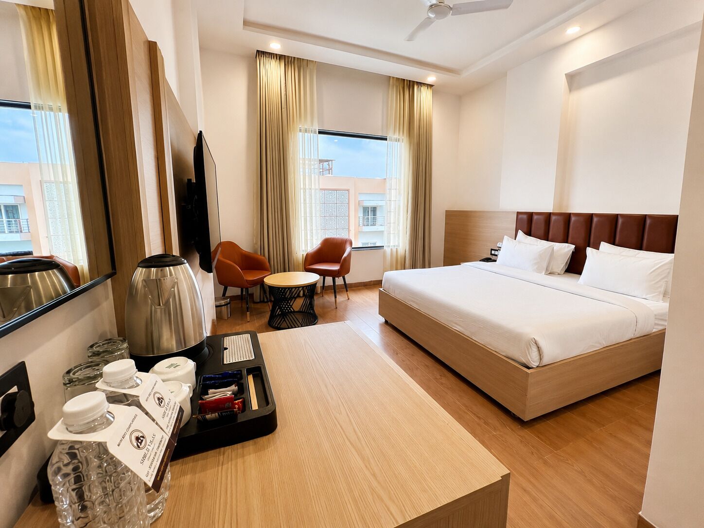 Quarto de hotel com cama, escrivaninha com cafeteira, uma cadeira e janela com cortinas.