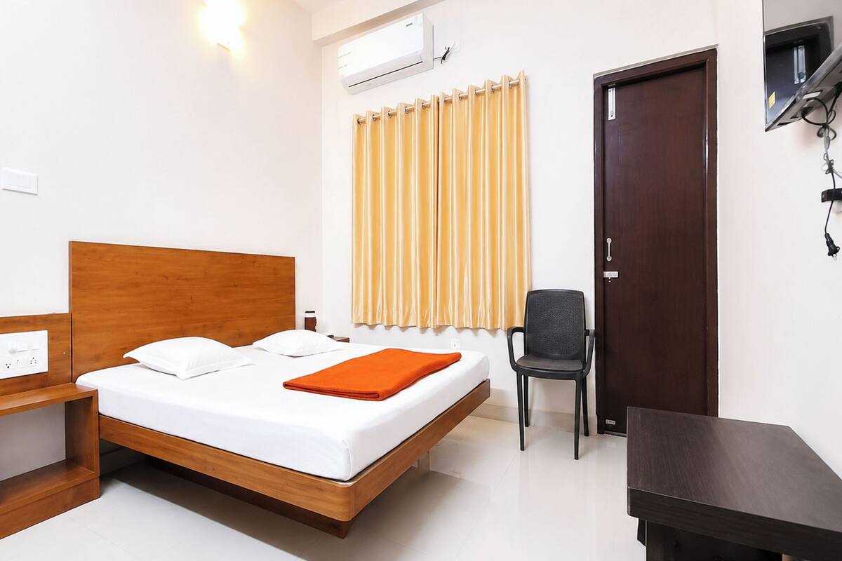 Quarto de hotel com cama de madeira, uma escrivaninha, uma cadeira, uma porta e um ar condicionado.
