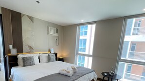 Quarto moderno com cama grande, mesas de cabeceira, uma mesinha e uma janela com vista para edifícios.