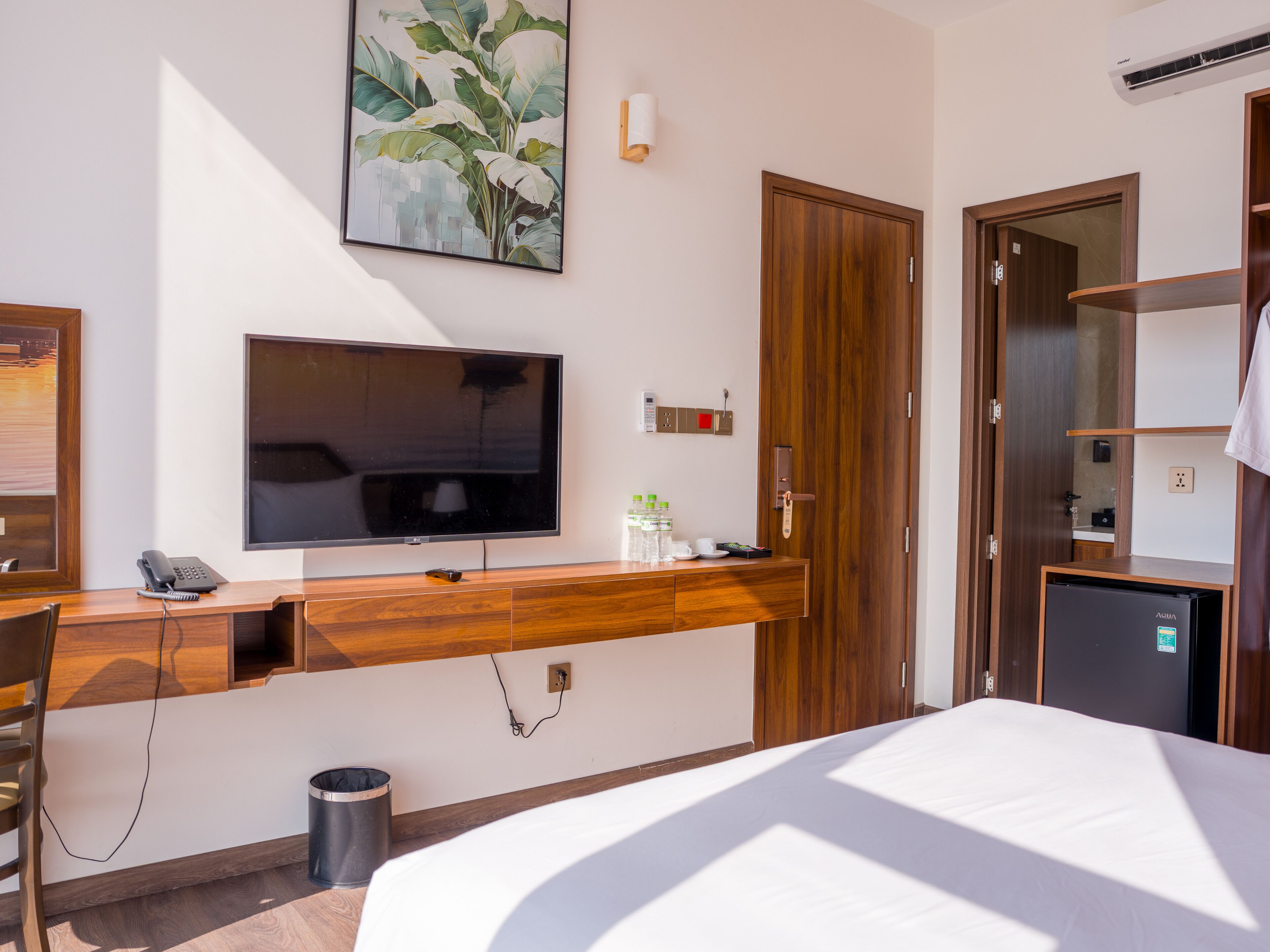 Quarto de hotel com cama, televisão na parede, secretária de madeira, lixeira e porta.