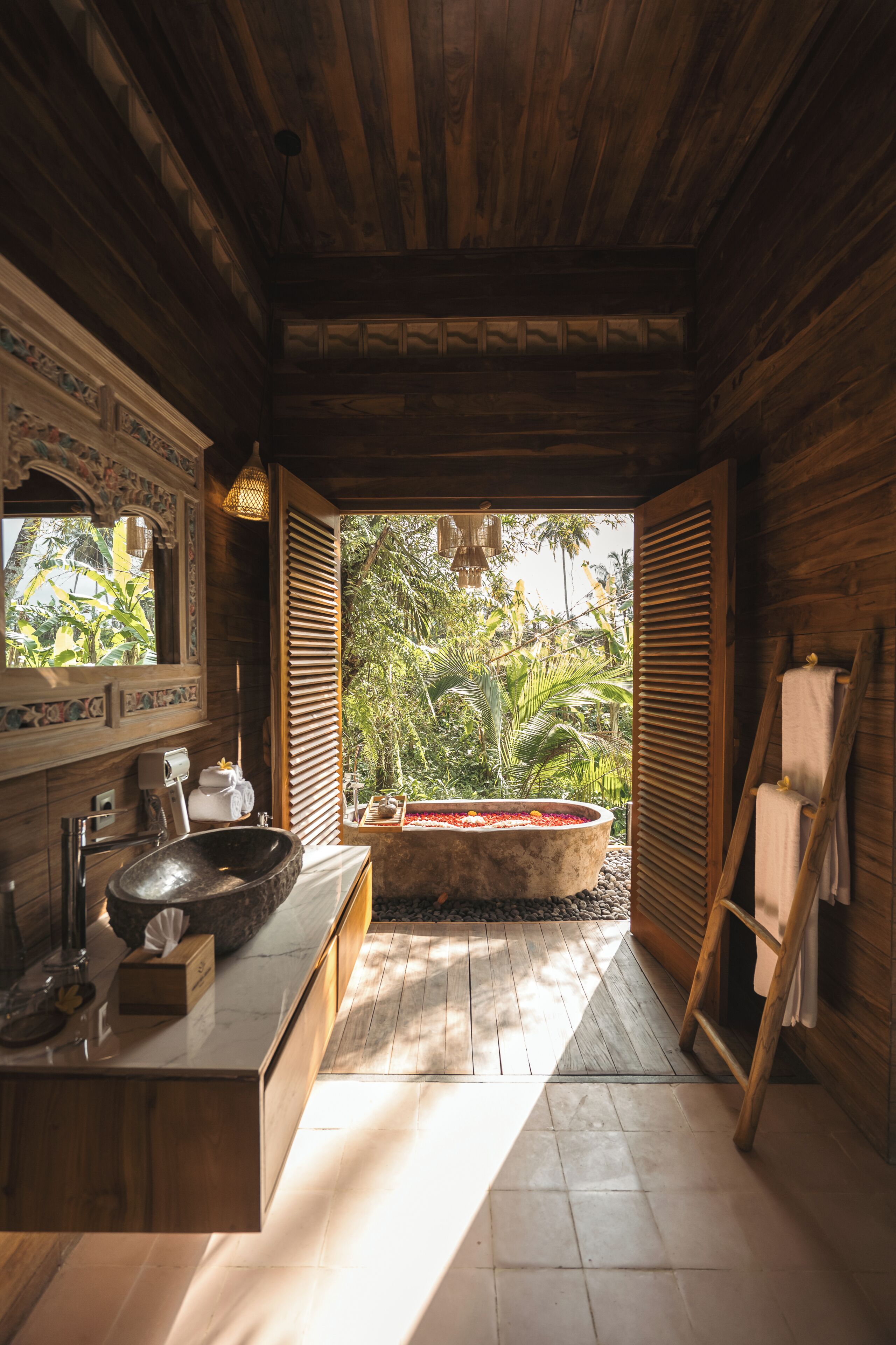 Interior em madeira com uma grande bacia de pedra, um banco de madeira e vista para vegetação tropical.