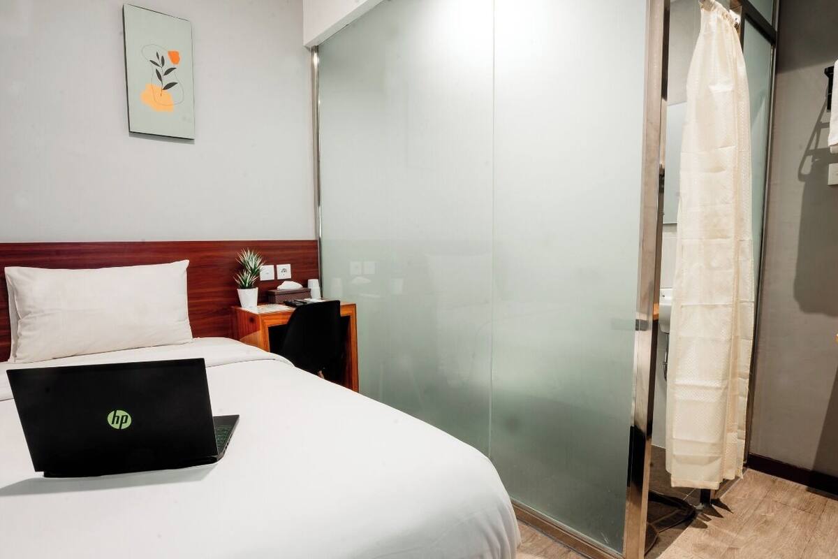 Quarto de hotel com cama, laptop, mesa de cabeceira de madeira, planta em vaso, box de vidro para chuveiro e porta-toalhas.