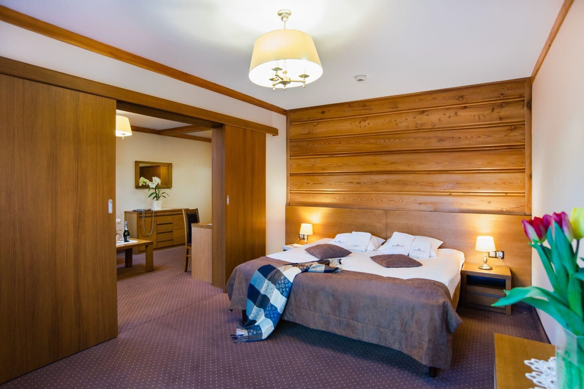Quarto de hotel com cabeceira de madeira, cama com manta xadrez, mesa de cabeceira com abajur, guarda-roupa de madeira e espelho.