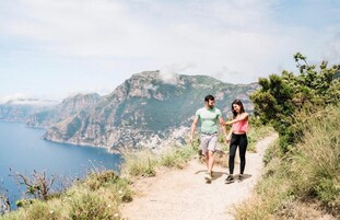 Duas pessoas caminham por uma trilha de terra com montanhas ao fundo e um corpo d'água.