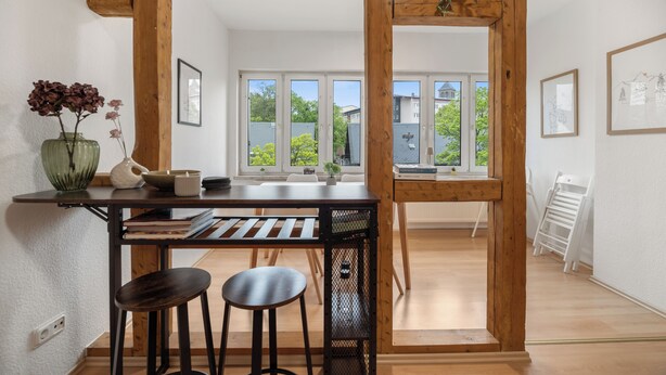 Uma área de refeições com uma mesa de madeira, dois banquinhos, um vaso com flores e quadros nas paredes.