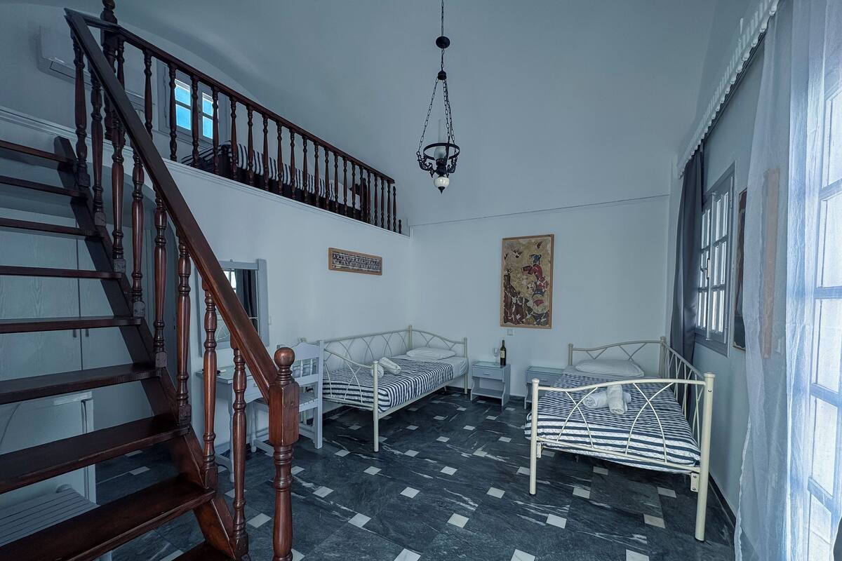 Quarto com escada de madeira, duas camas com roupa de cama listrada, luminária pendente e uma grande janela com cortinas.