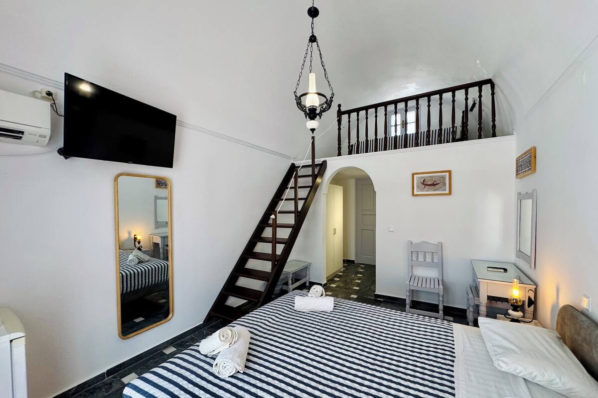 Quarto de hotel com cama, espelho, televisão, escada e uma pequena mesa com abajur.