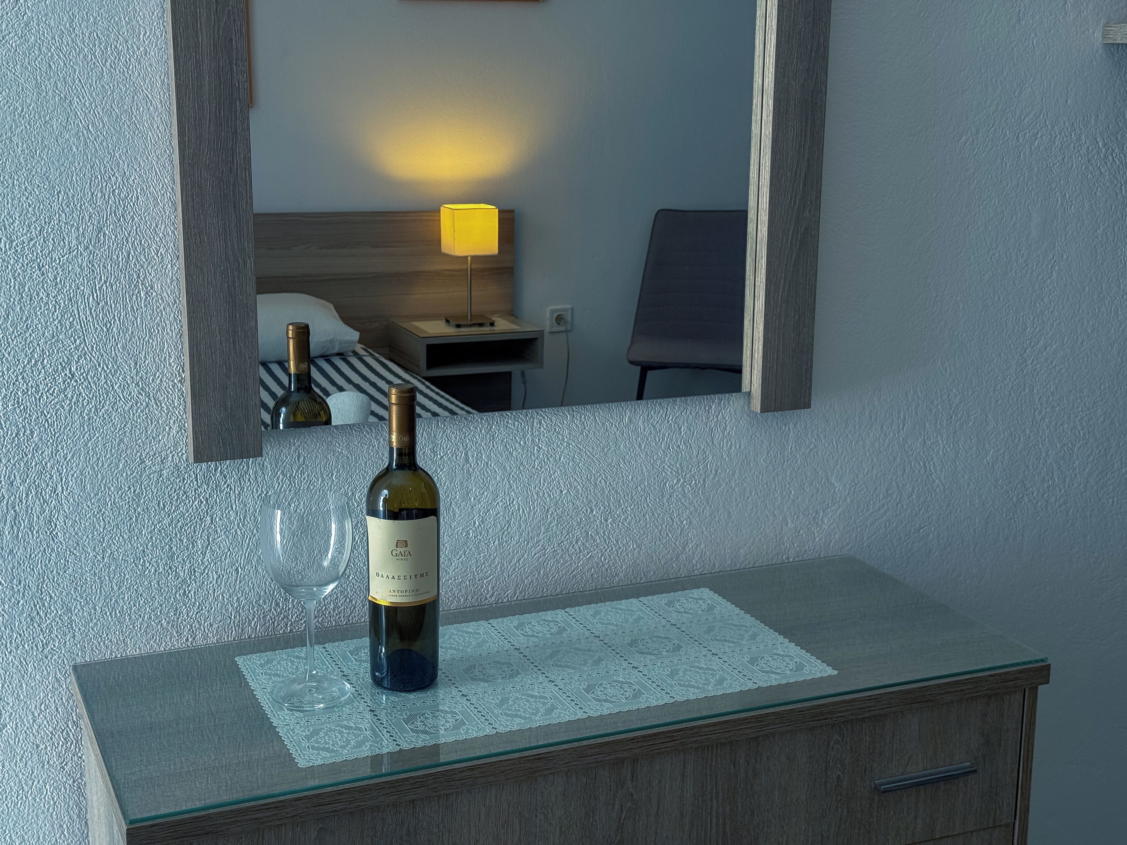 Quarto de hotel com bancada de vidro, uma garrafa de vinho e uma taça de vinho no balcão.