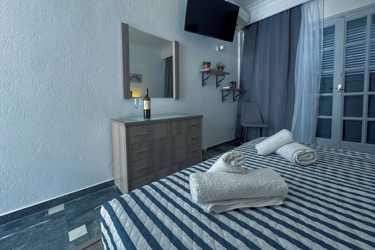 Quarto de hotel com cama, televisão fixada na parede, penteadeira, espelho, janela com cortinas e uma mesinha com uma planta.
