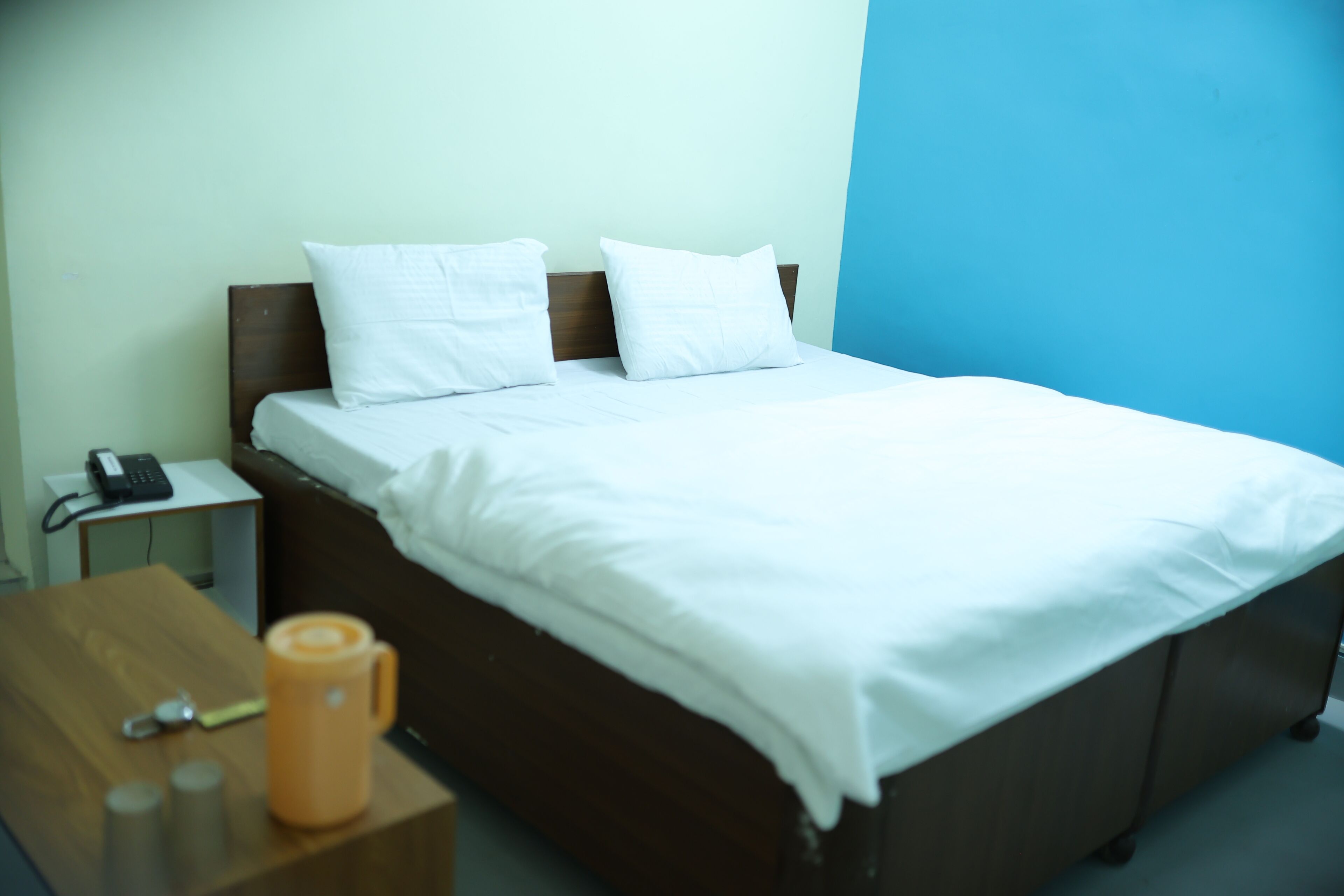 Uma cama de solteiro com roupa de cama branca, cabeceira de madeira, mesa de cabeceira com um telefone e uma caneca, e parede azul.
