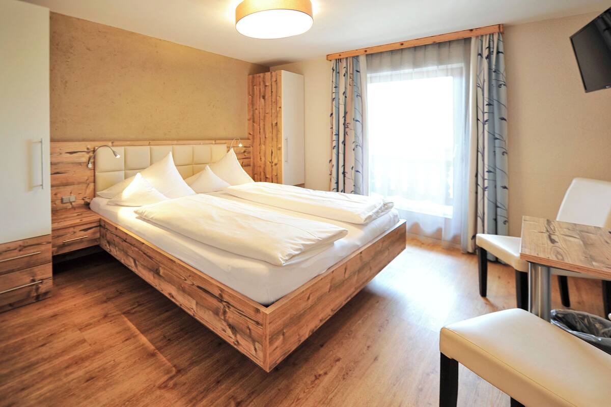 Uma cama de madeira com roupa de cama branca, uma mesa de cabeceira de madeira, uma escrivaninha com cadeira e uma televisão na parede.