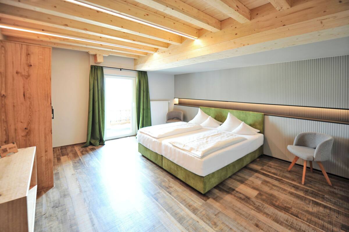 Quarto moderno com teto de madeira, cama estofada em verde, mesa de cabeceira e uma cadeira.
