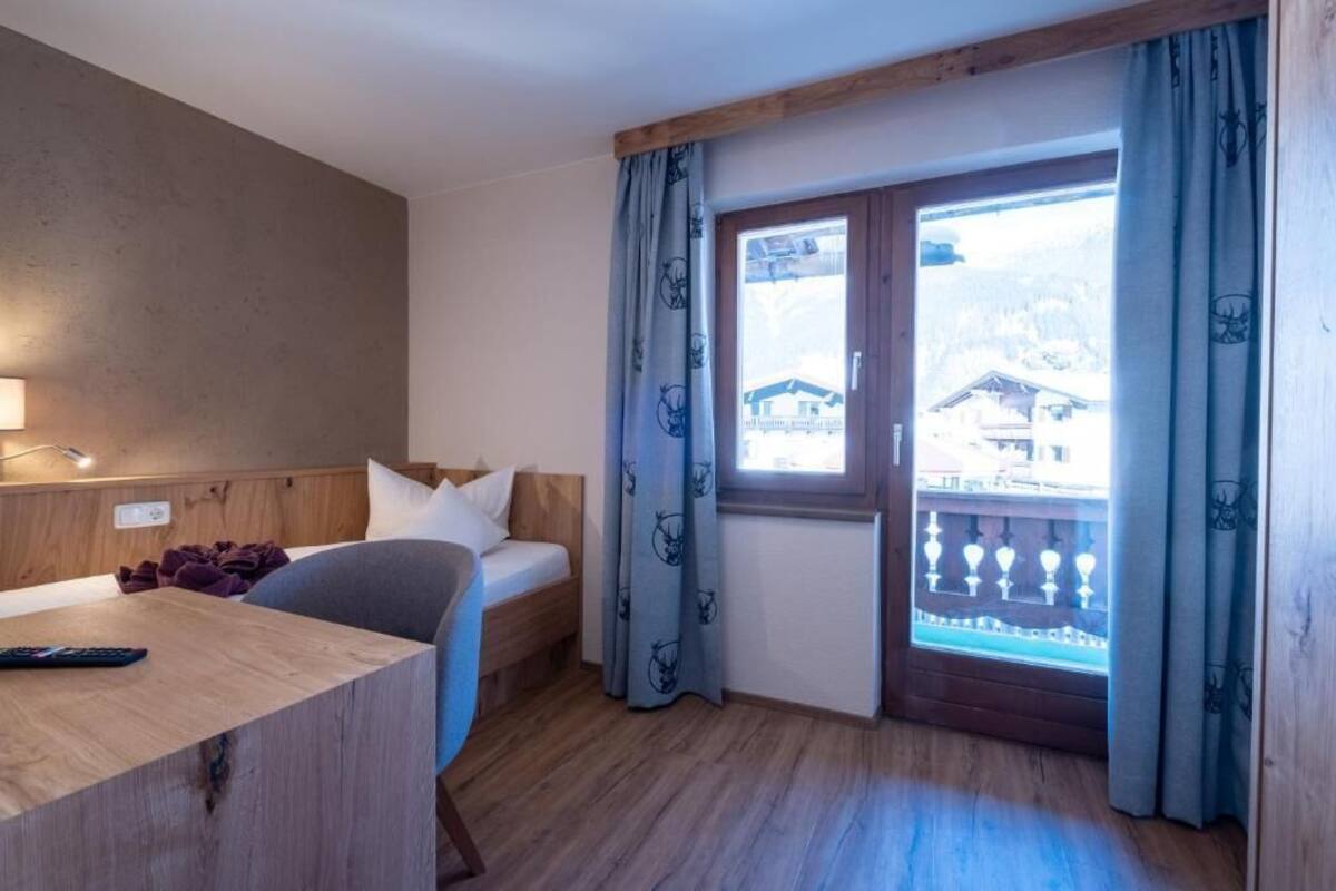 Quarto de hotel com cama de madeira, uma escrivaninha, uma cadeira, janela com cortinas e vista para uma montanha.