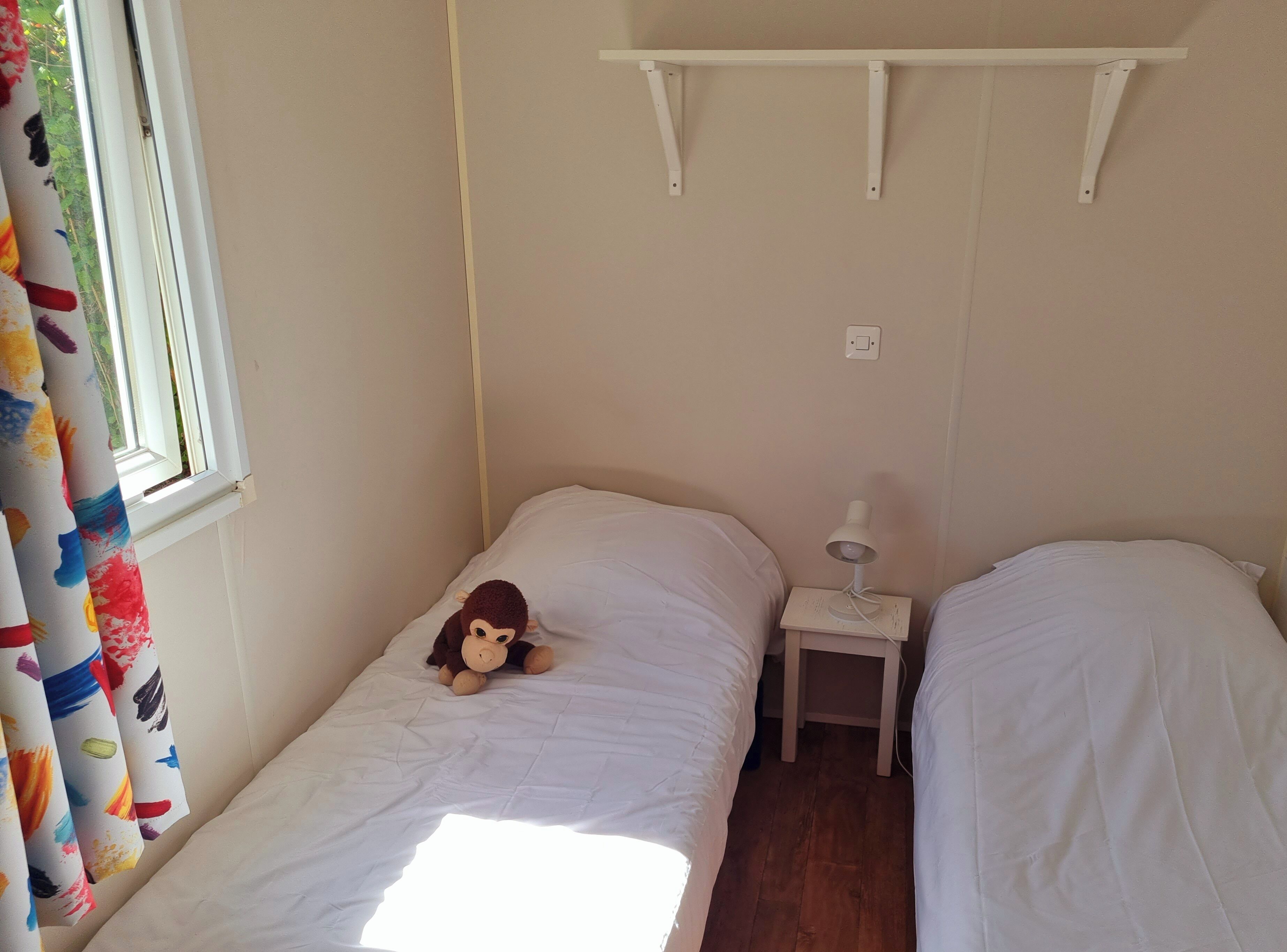 Uma cama de solteiro com uma pequena mesa de cabeceira e uma luminária, uma janela com cortinas, e uma parede branca.