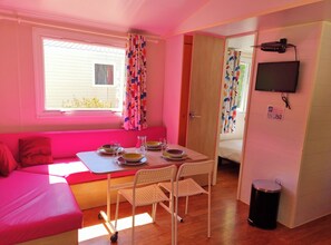 Quarto com sofá rosa, mesa de jantar com cadeiras, TV fixada na parede e janela com cortinas.