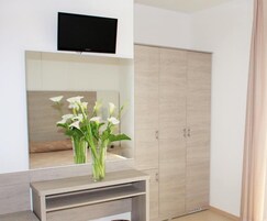 Quarto com televisão na parede, espelho refletindo um vaso de flores brancas, guarda-roupa e mesa de apoio.