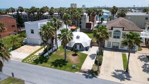 Zona residencial com um edifício branco em forma de cúpula, cercado por palmeiras, adjacente a uma rua com carros estacionados e um prédio com telhado marrom.