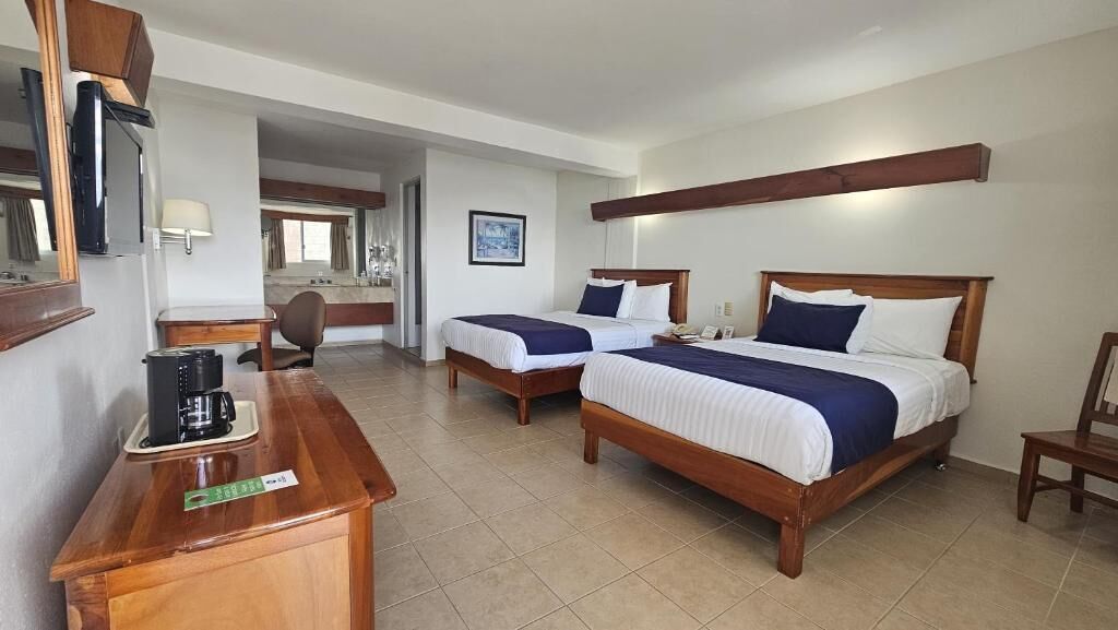 Quarto de hotel com duas camas, uma mesa de trabalho com cafeteira, televisão e um quadro na parede.