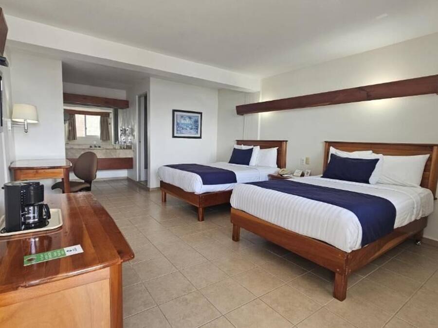 Quarto de hotel com duas camas, uma mesa de trabalho com cafeteira, televisão e um quadro na parede.