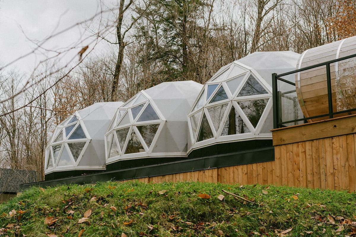 Uma série de estruturas em forma de cúpula geodésica com grandes janelas, paredes de madeira e um primeiro plano gramado.