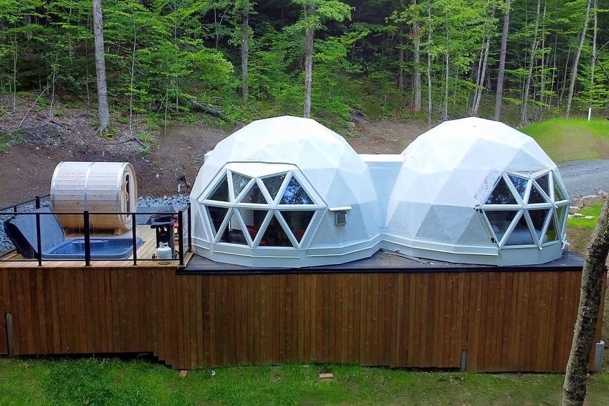 Duas estruturas em forma de cúpula geodésica com um deck de madeira à frente, rodeadas por uma floresta.