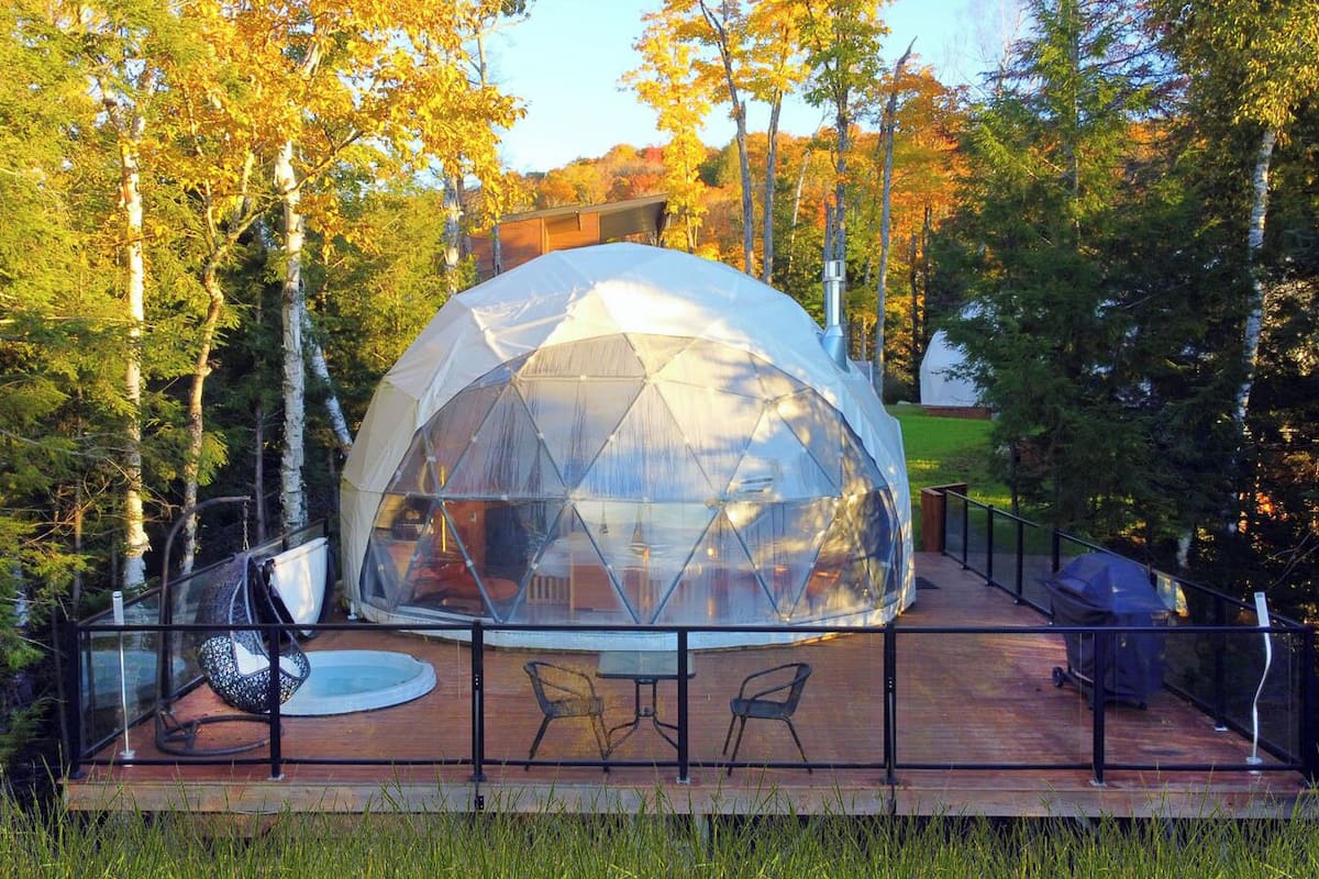 Uma estrutura em forma de cúpula geodésica com fachada de vidro, rodeada por um deck de madeira e guarda-corpos de metal.