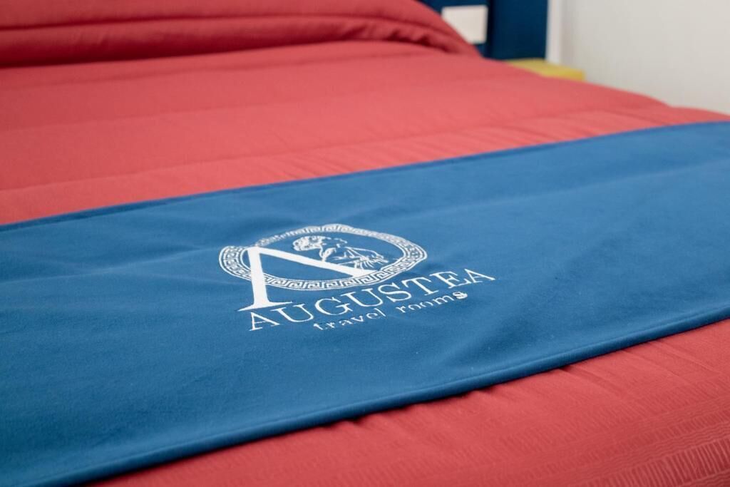 Uma cama com colcha azul e vermelha exibindo o logotipo e o nome 'Auguste Tea' e o texto 'travel rooms'.