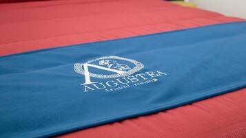 Uma cama com colcha azul e vermelha exibindo o logotipo e o nome 'Auguste Tea' e o texto 'travel rooms'.
