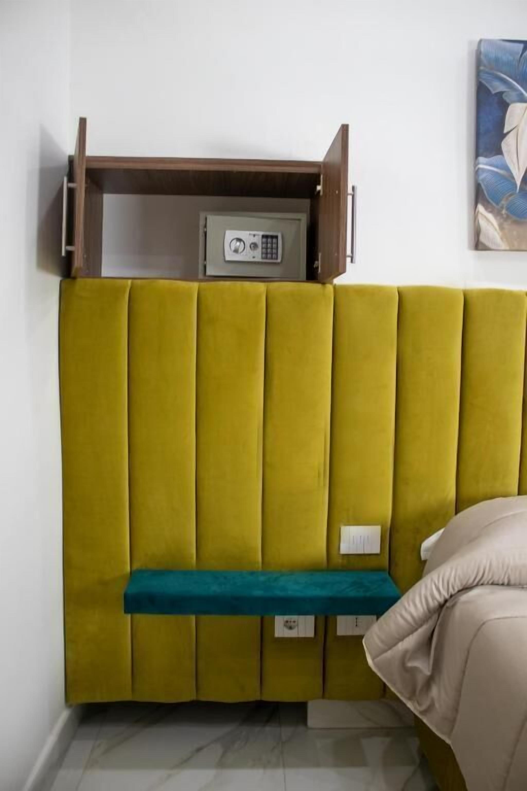 Quarto com cabeceira estofada amarela, um banquinho azul-turquesa, mesa de cabeceira branca e rádio fixado na parede.