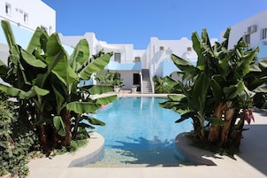 Uma piscina cercada por edifícios brancos, com grandes bananeiras e um céu azul claro.